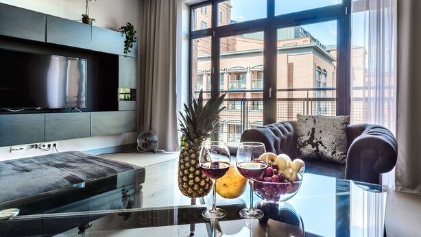 Uma sala de estar moderna com uma janela ampla, uma TV de tela plana, uma mesa de centro de vidro com frutas e taças de vinho, e um sofá de cor escura.