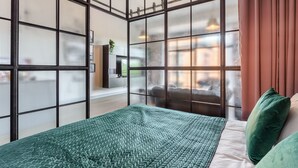 Quarto de hotel moderno com uma cama grande, colcha verde texturizada e vista através de uma janela ampla com grade de metal.