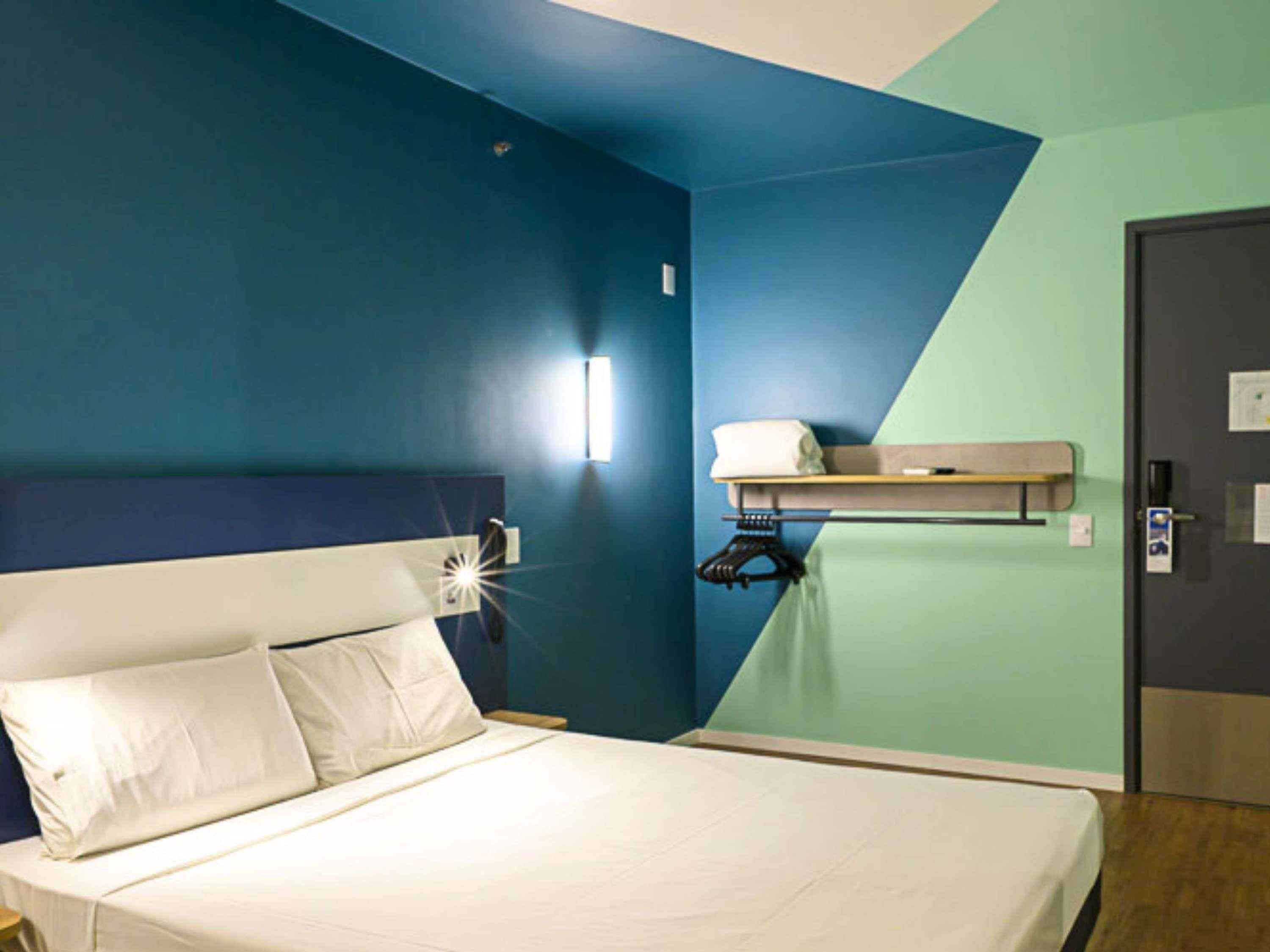 Quarto de hotel com uma cama, uma prateleira na parede com uma toalha e uma porta com fechadura.