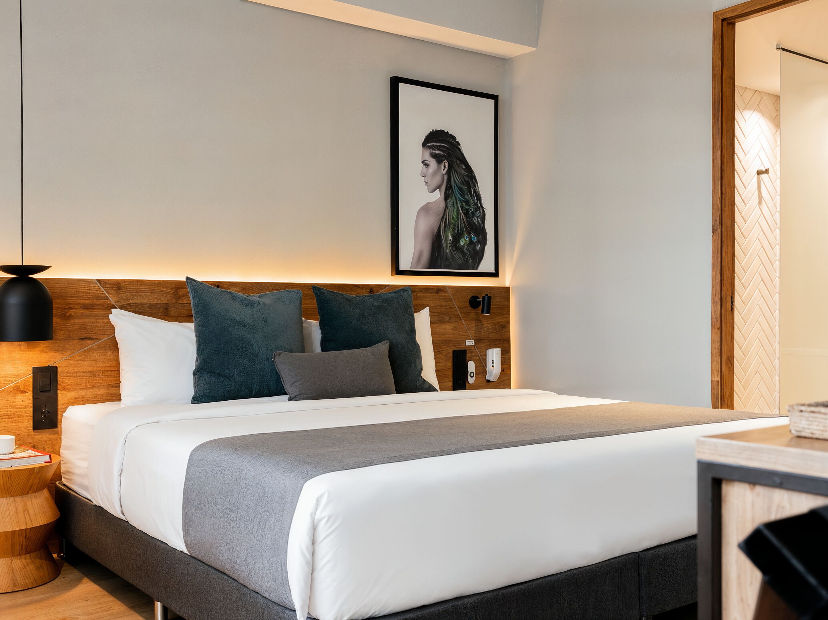 Quarto de hotel moderno com cabeceira de madeira, cama com roupa de cama branca e cinza, uma obra de arte emoldurada sobre a cama, mesa de cabeceira com abajur e criado-mudo de madeira.