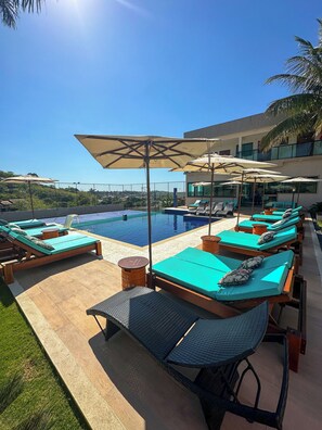 Área à beira da piscina com espreguiçadeiras e guarda-sóis, com vista clara para a piscina e um edifício ao fundo.
