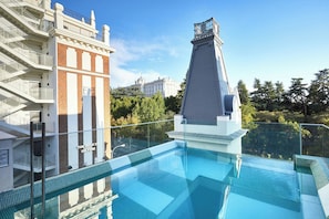 Uma piscina infinita no terraço, com guarda-corpo de vidro, um edifício branco com fachada de tijolos e um céu azul límpido.