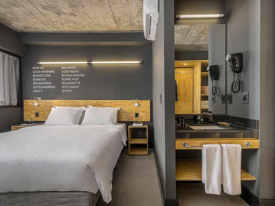 Quarto de hotel moderno com cama grande, mesas de cabeceira, cabeceira de madeira, secretária embutida com espelho e banheiro com pia e porta-toalhas.
