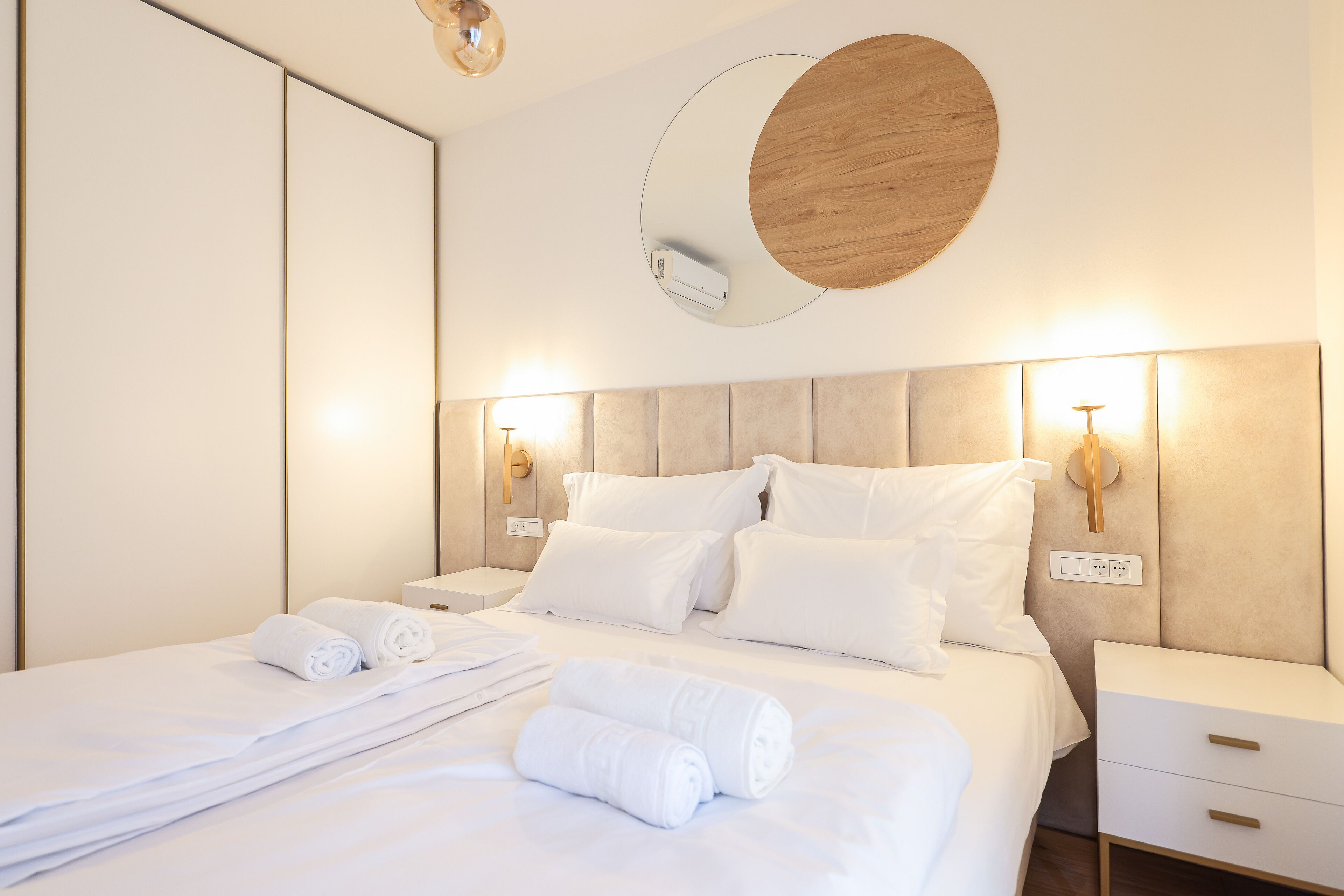 Quarto de hotel moderno com uma cama bem arrumada, duas mesas de cabeceira e um espelho redondo grande na parede.