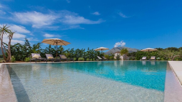 Uma piscina cristalina com espreguiçadeiras e guarda-sóis, cercada por vegetação tropical e uma montanha ao fundo.
