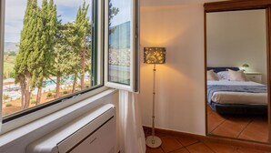 Quarto com cama, mesa de cabeceira, abajur e vista para árvores e piscina lá fora.
