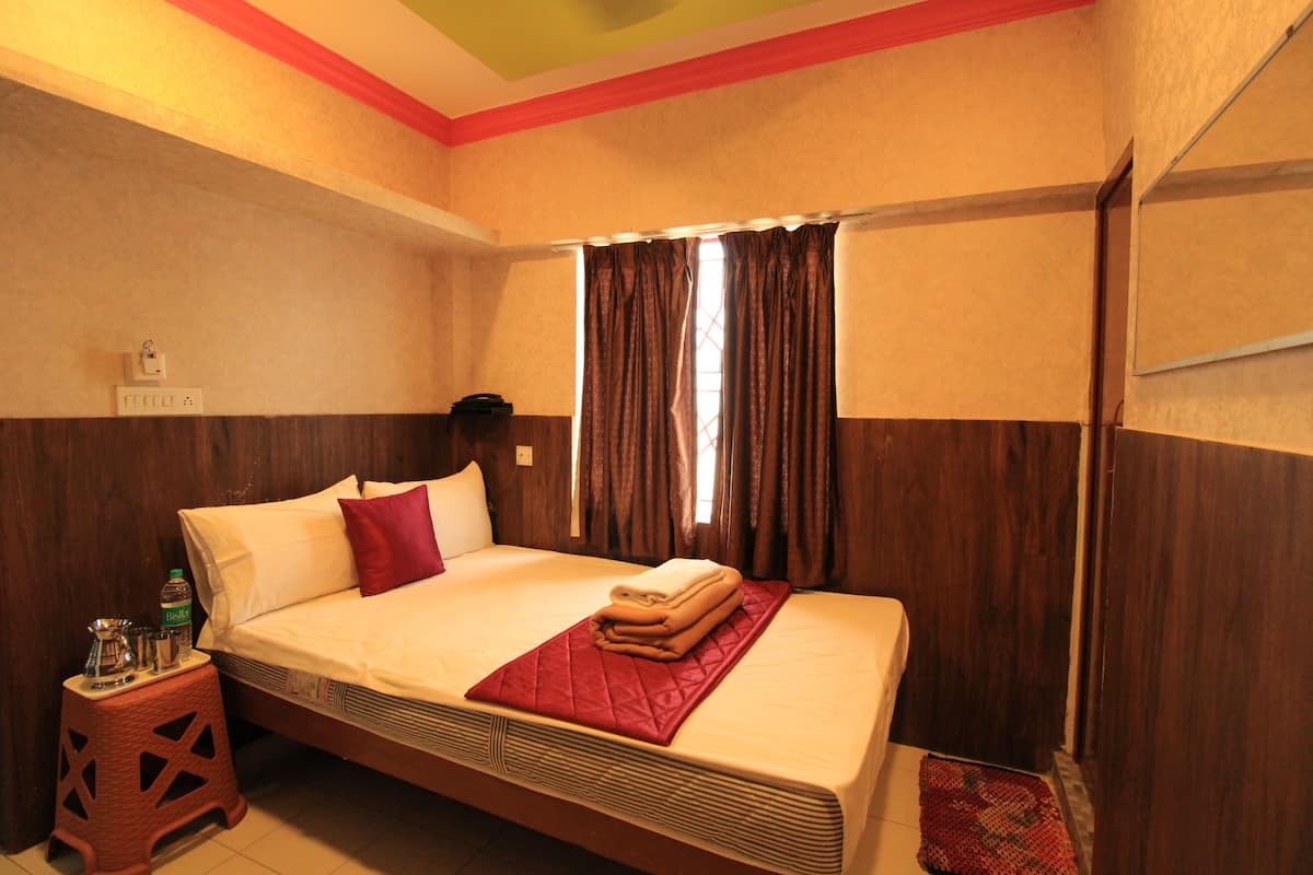 Uma cama bem arrumada com um cobertor vermelho, uma mesa de cabeceira de madeira com uma garrafa e copos, e uma janela com cortinas.