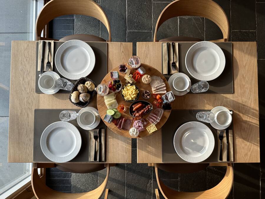 Mesa de jantar posta para quatro pessoas, com diversos alimentos e bebidas, rodeada por cadeiras de madeira.