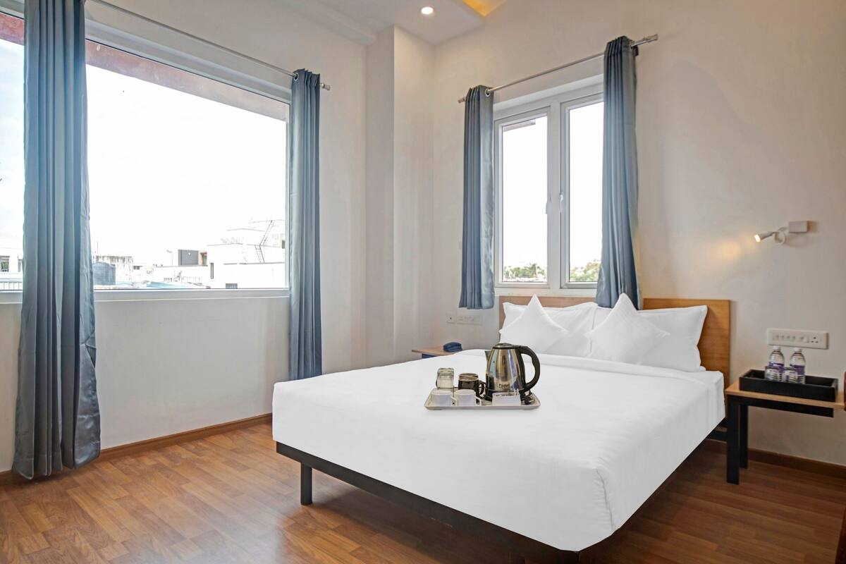 Quarto de hotel com uma cama grande, uma mesa de cabeceira com chaleira, um tabuleiro com xícaras e um termo, e uma janela com cortinas.