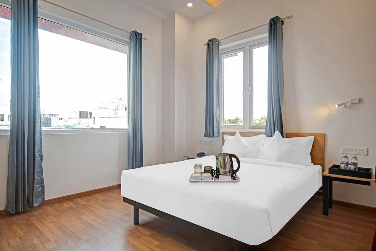 Quarto de hotel com uma cama grande, uma mesa de cabeceira com chaleira e xícaras, e uma janela com cortinas.