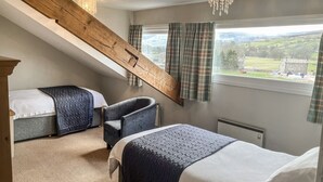 Quarto com teto inclinado, duas camas, uma cadeira, um lustre e vista para o campo.
