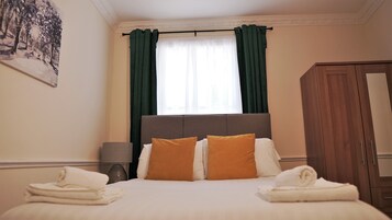 Quarto de hotel com cama, duas almofadas, mesa de cabeceira, luminária, guarda-roupa e janela com cortinas.