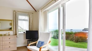 Quarto com televisão, cadeira com almofada, cómoda e porta de correr para área externa com vista para o mar.