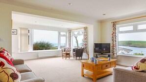 Uma sala de estar espaçosa com uma grande janela que oferece vista para o mar, uma mesa de centro de madeira e uma televisão.