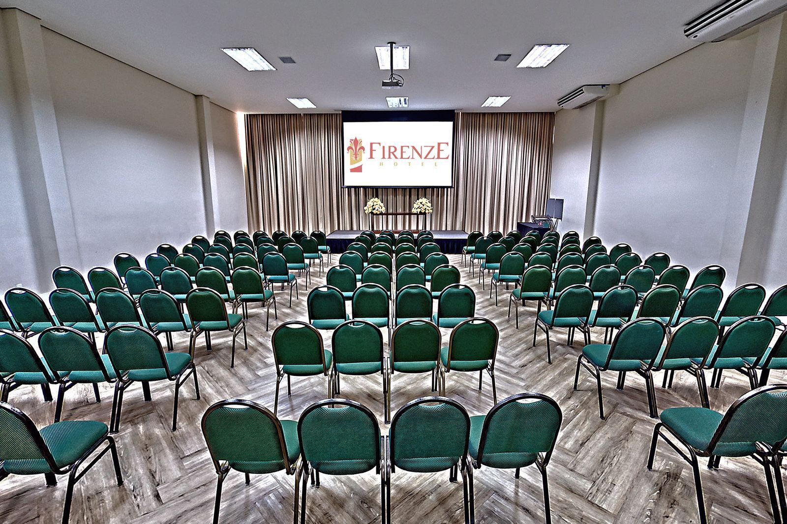 Uma sala de conferências com fileiras de cadeiras verdes voltadas para um palco com uma tela exibindo 'FIRENZE HOTEL'.