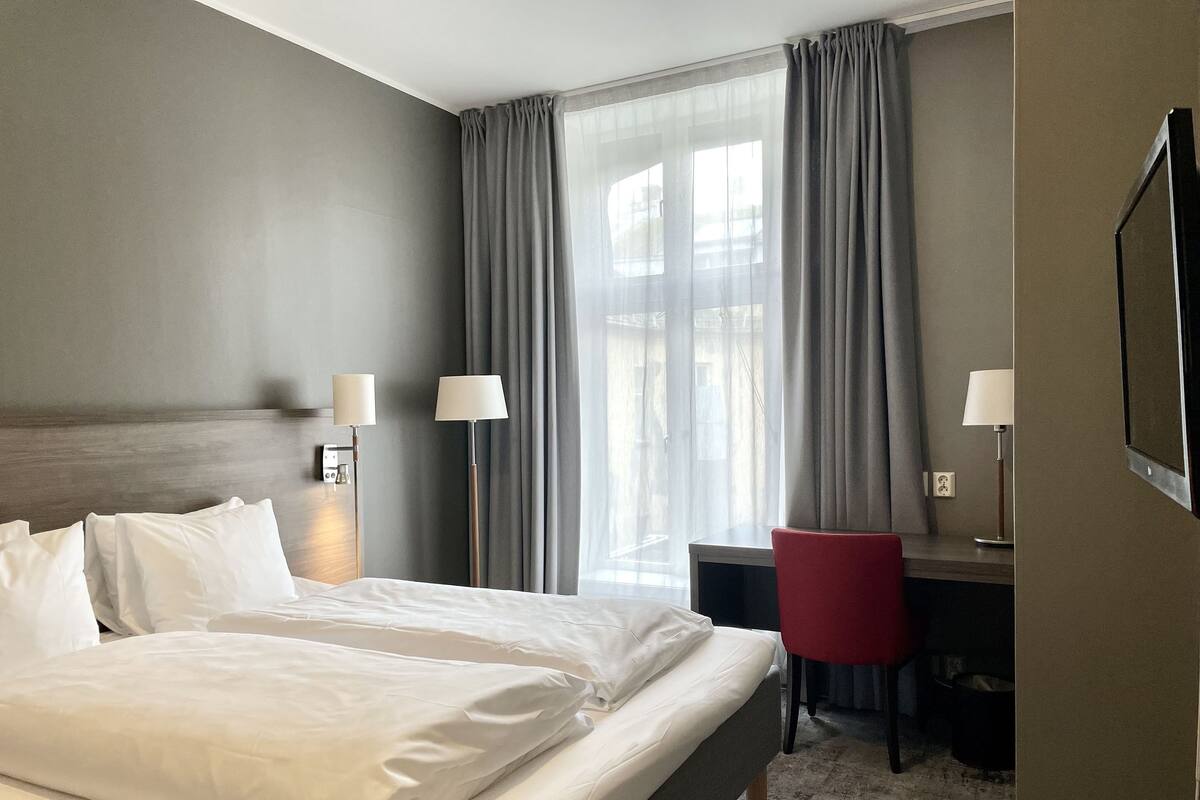 Quarto de hotel com uma cama grande, duas luminárias de cabeceira, uma mesa com cadeira e uma janela ampla com cortinas.