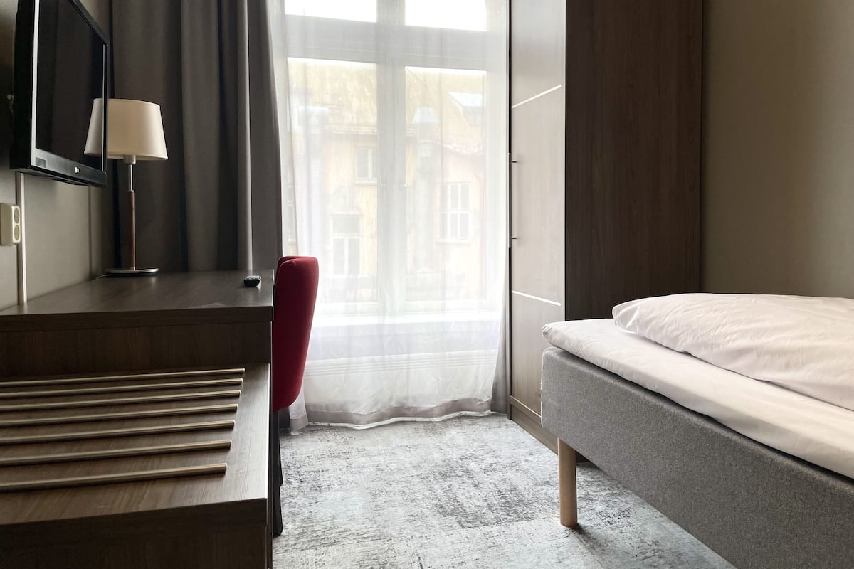 Quarto de hotel moderno com escrivaninha de madeira, televisão, cama com colchão cinza e vista da cidade através de cortinas leves.