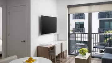 Um apartamento moderno com televisão de tela plana na parede, sala de jantar com mesa e cadeiras, e varanda com vista para o exterior do edifício.