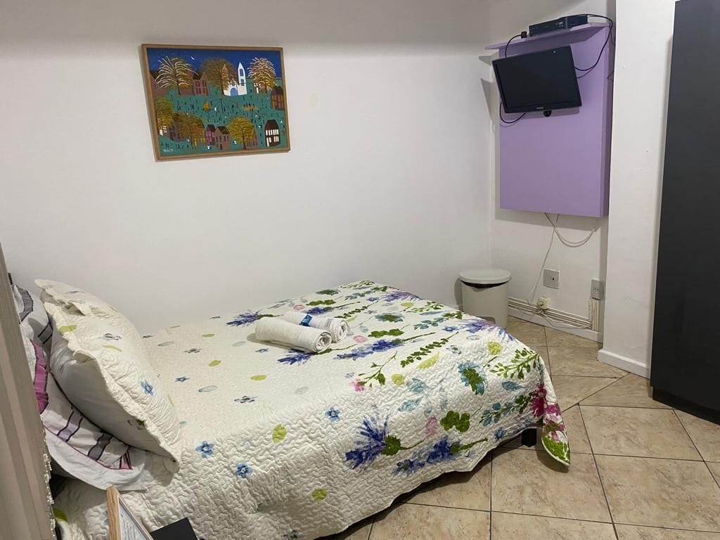 Quarto com cama, um cesto de lixo pequeno, televisão na parede e um quadro na parede.