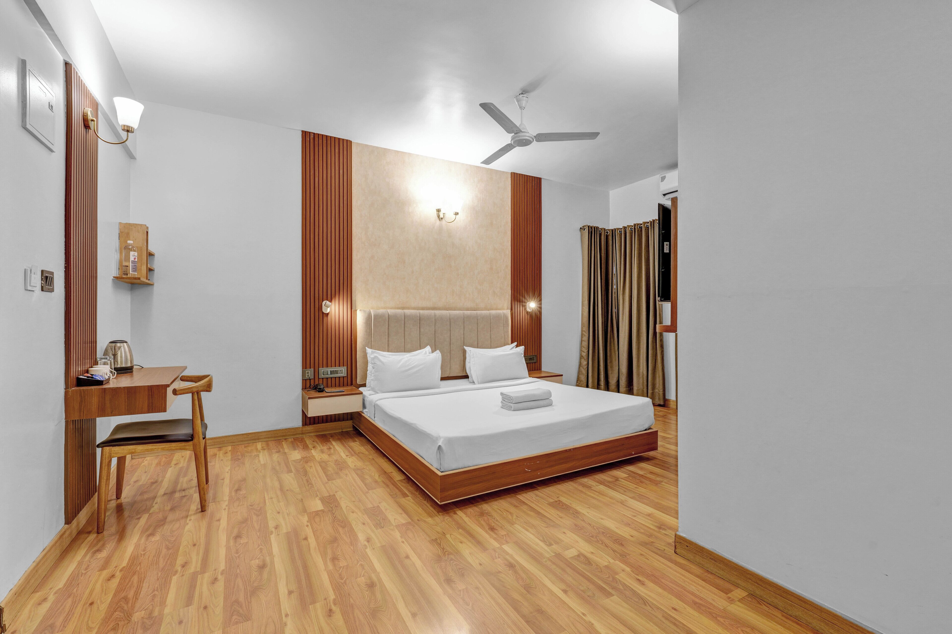 Quarto de hotel moderno com piso de madeira, cama com lençóis brancos, escrivaninha com cadeira e ventilador de teto.