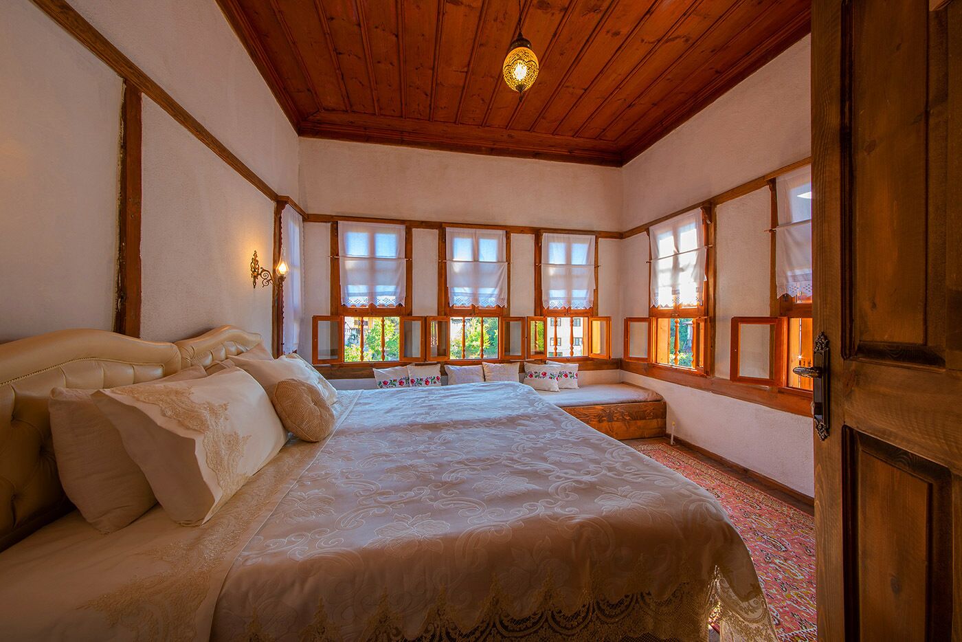Quarto com cama grande, um banco e várias janelas com cortinas.