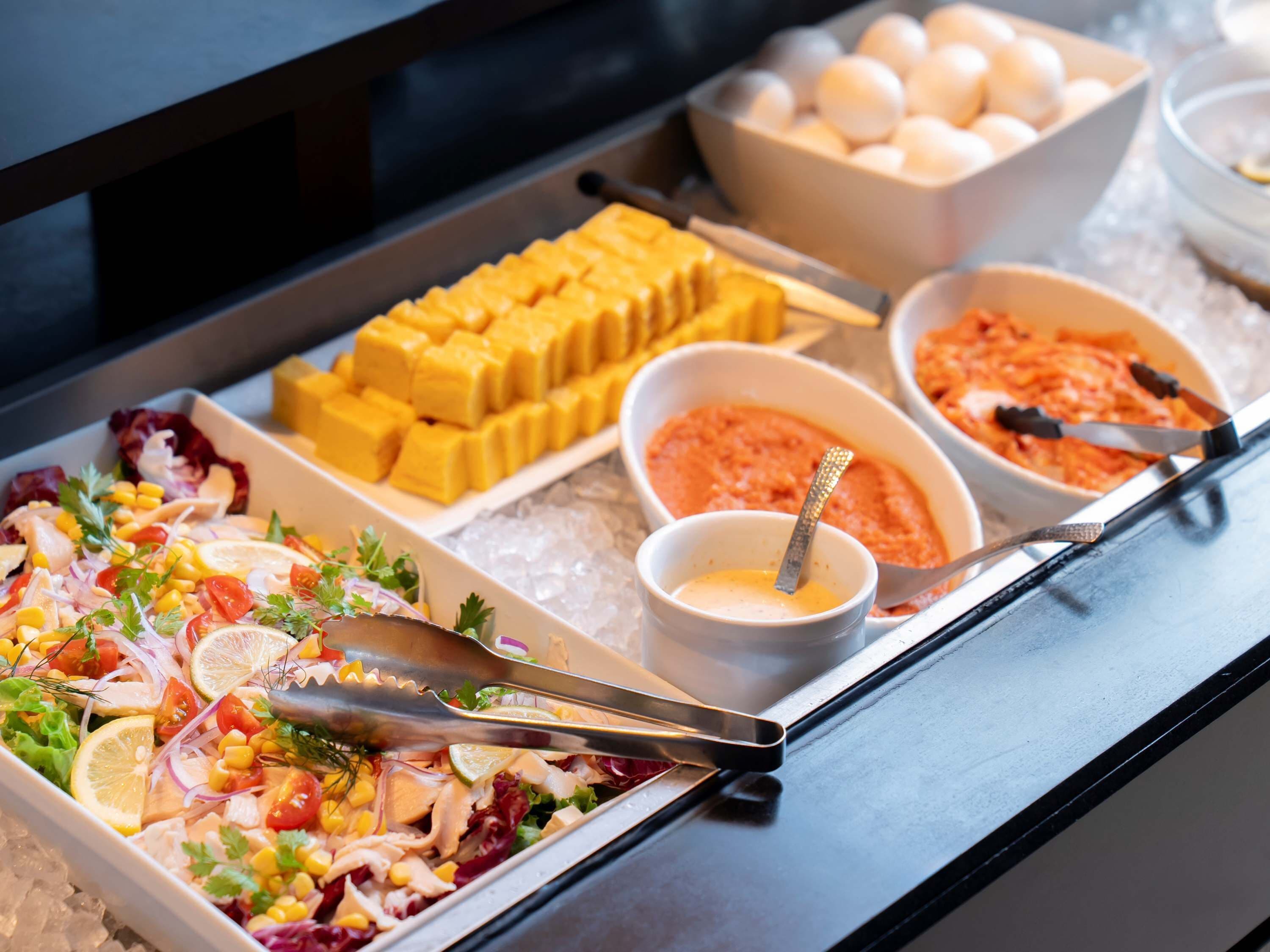 Buffet com diversos alimentos, incluindo salada, cubos de queijo, ovos e molhos.