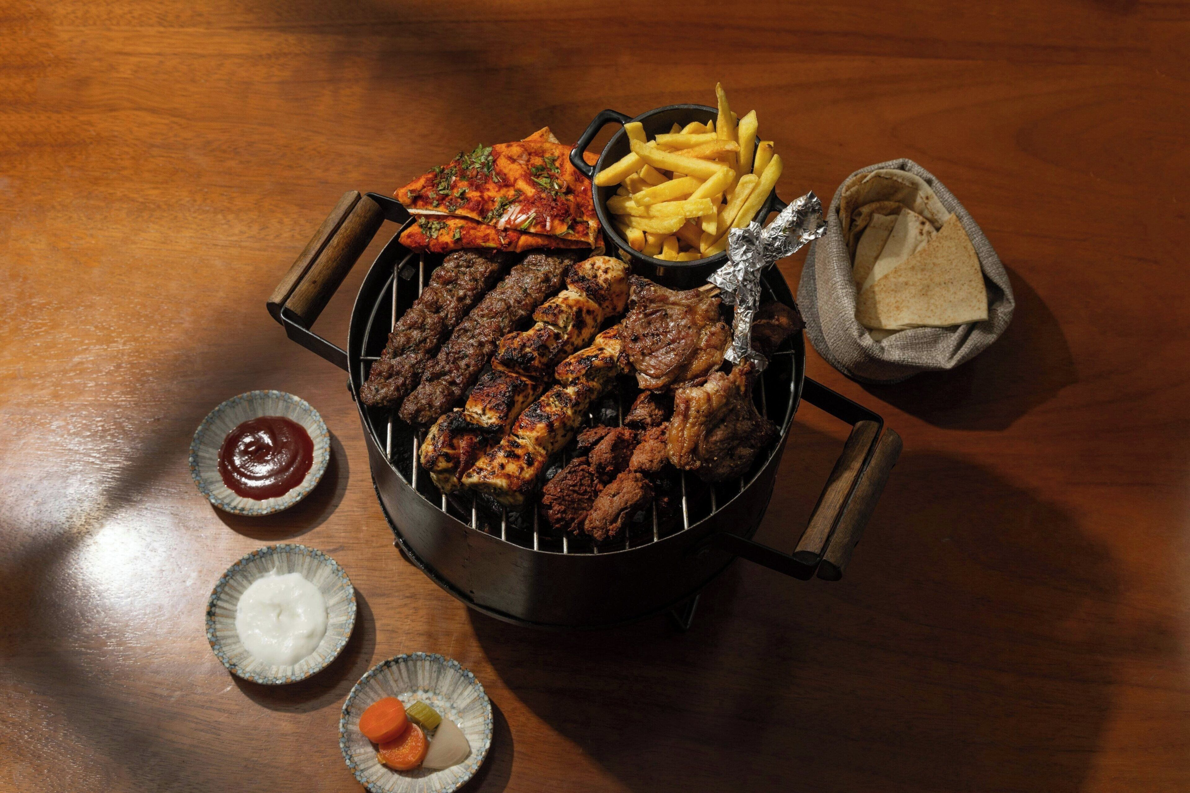 Uma churrasqueira com diversos tipos de carne e acompanhamentos, incluindo batatas fritas, pão sírio e molhos.