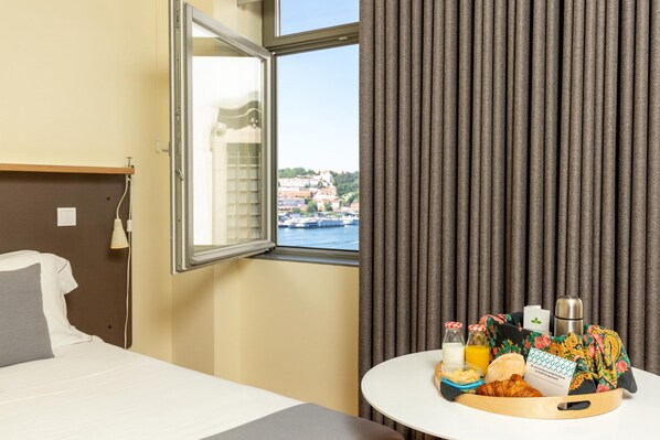 Quarto de hotel com cama, mesa de cabeceira com itens de café da manhã, janela com vista para prédios e cortina.