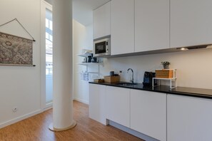 Uma cozinha moderna com armários brancos, bancada preta e um micro-ondas. O chão é de madeira e há uma janela que ilumina naturalmente o espaço.