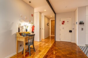 Um corredor com piso de madeira, uma mesa com cadeira, um extintor de incêndio e uma porta com sinalização de saída de emergência.
