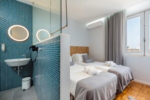 Quarto de hotel com banheiro com azulejos azuis em mosaico, pia e espelho. O quarto tem duas camas com roupa de cama cinza e uma janela com cortinas.