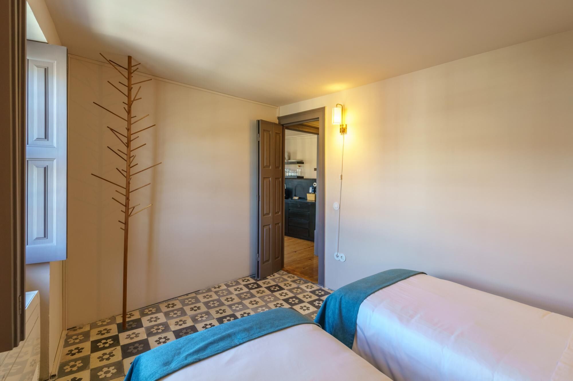 Quarto de hotel com duas camas, piso estampado, um objeto decorativo de madeira e cozinha visível por uma porta aberta.