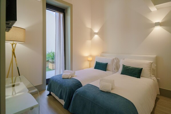 Quarto de hotel com duas camas, uma mesinha de cabeceira, uma luminária e vista para o exterior.
