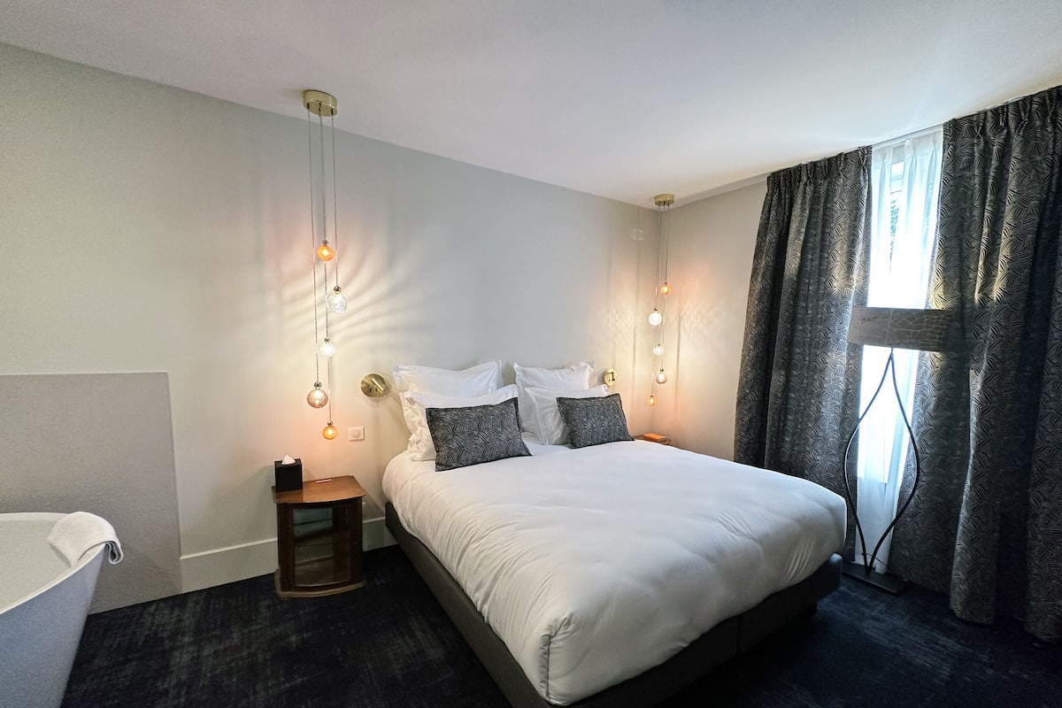 Quarto de hotel moderno com uma cama grande, uma mesa de cabeceira, uma luminária na parede e uma janela com cortinas.
