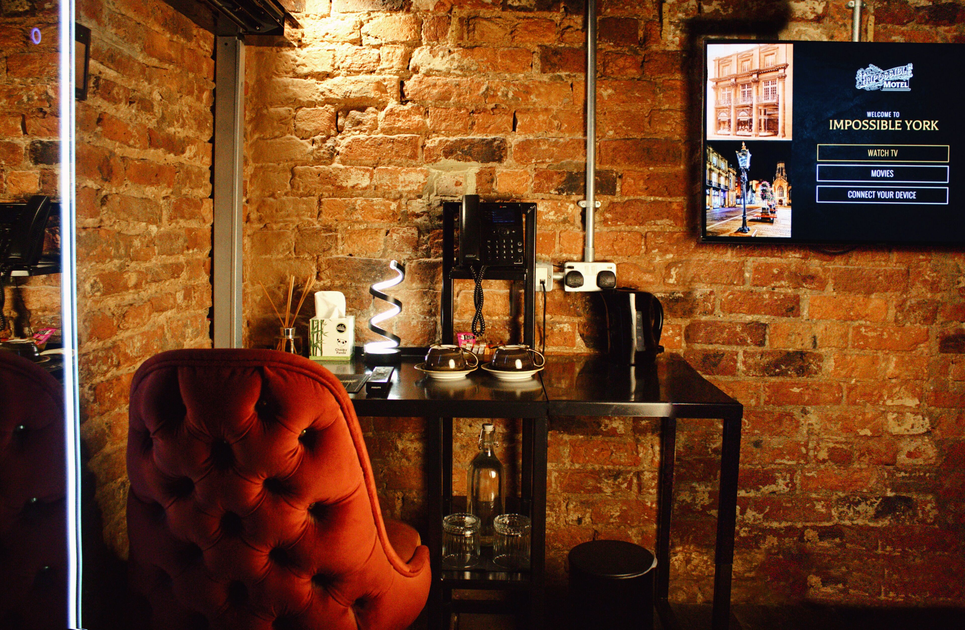 Quarto com paredes de tijolo, uma poltrona estofada em vermelho, uma máquina de café e uma TV exibindo "Welcome to IMPOSSIBLE YORK" com opções para assistir TV, filmes e conectar um dispositivo.