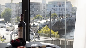 Uma mesa com uma garrafa de vinho, dois copos e uma cesta de frutas em um quarto com vista para uma ponte e edifícios.