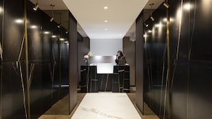 Corredor moderno com piso de mármore e paredes escuras, que dá acesso a uma área de recepção com balcão e uma pessoa em pé atrás dele.