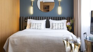 Quarto de hotel com uma cama grande, abajures, um espelho redondo e uma mesa de jantar com um copo de vinho e frutas.