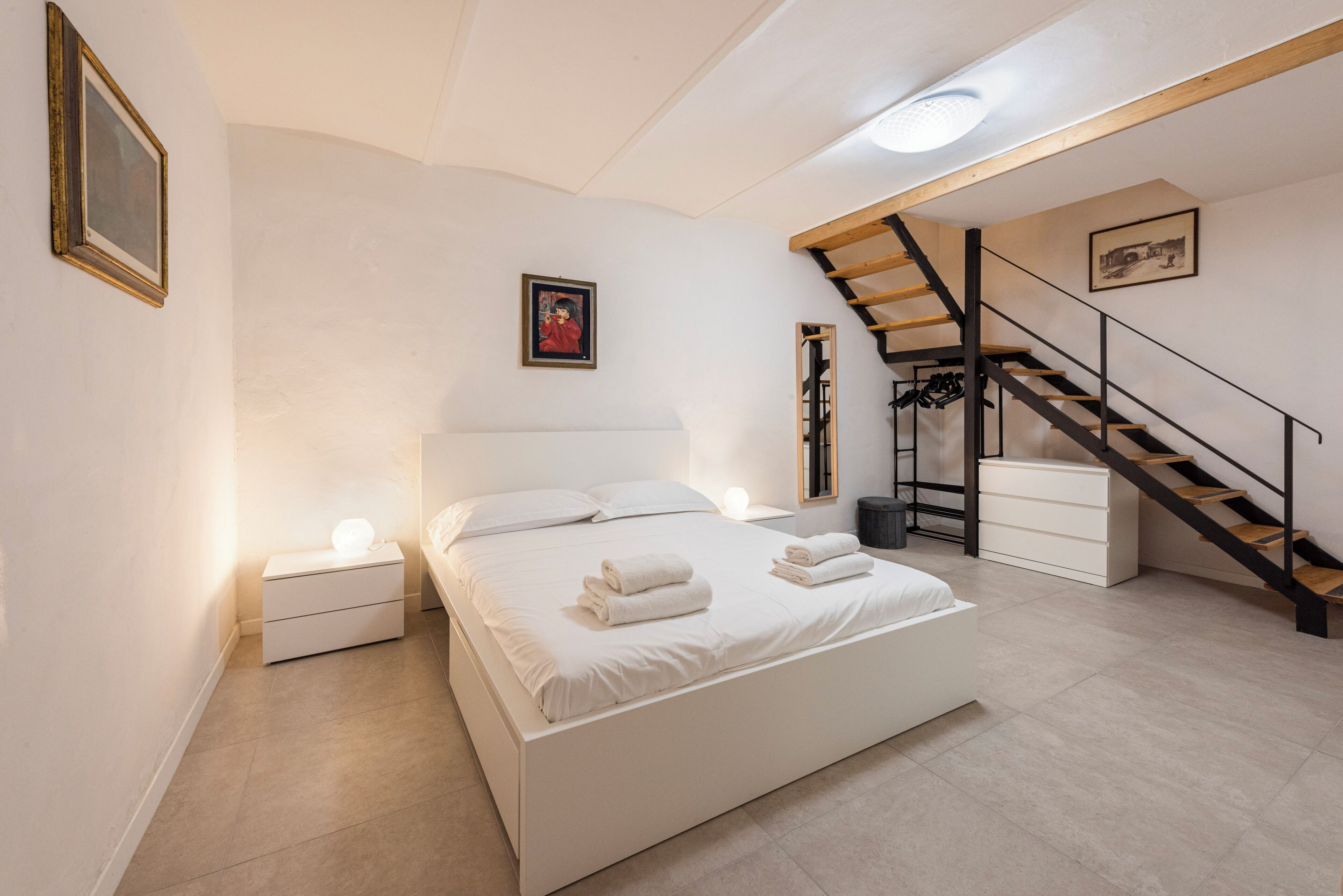 Quarto com cama grande, mesa de cabeceira, cômoda, escada e quadro na parede.