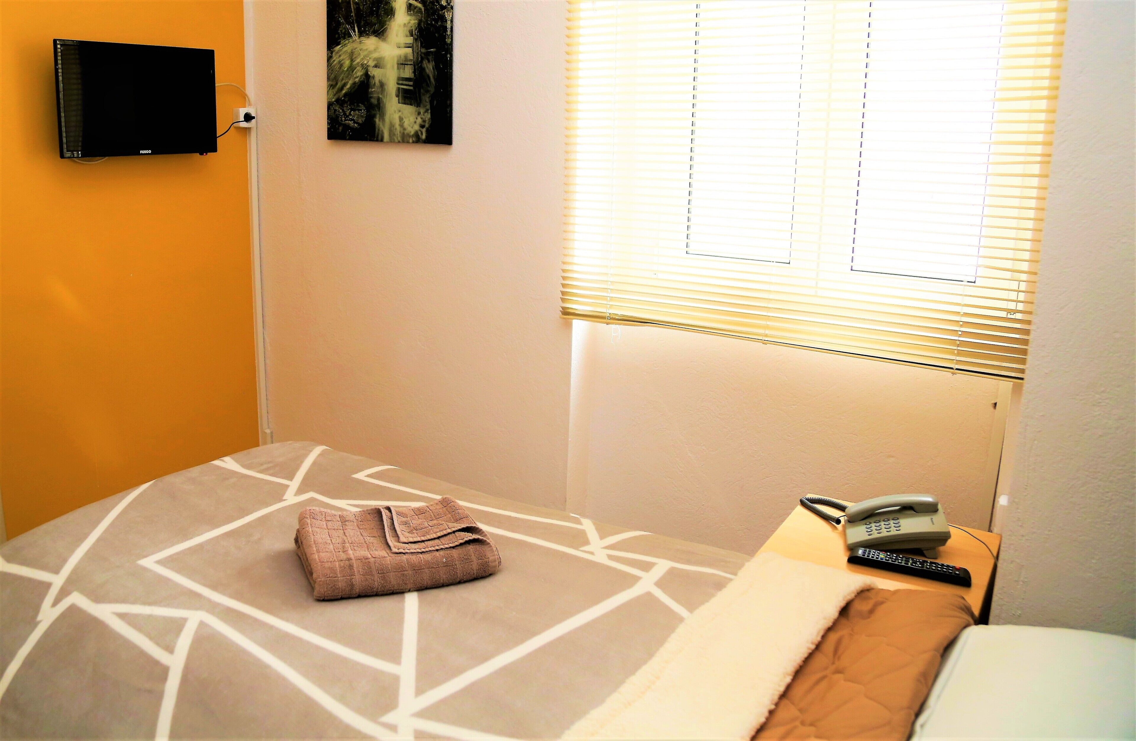 Quarto de hotel com cama, mesa de cabeceira com telefone e controle remoto, televisão na parede e janela com persianas.