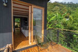 Uma casa moderna com deck de madeira, porta de vidro que dá para uma sala de estar com sofá e poltronas, e vista para uma floresta verde exuberante.