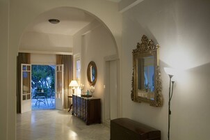 Um corredor com um banco de madeira, um espelho e um candeeiro. Ao fundo, uma mesa com um candeeiro e velas.