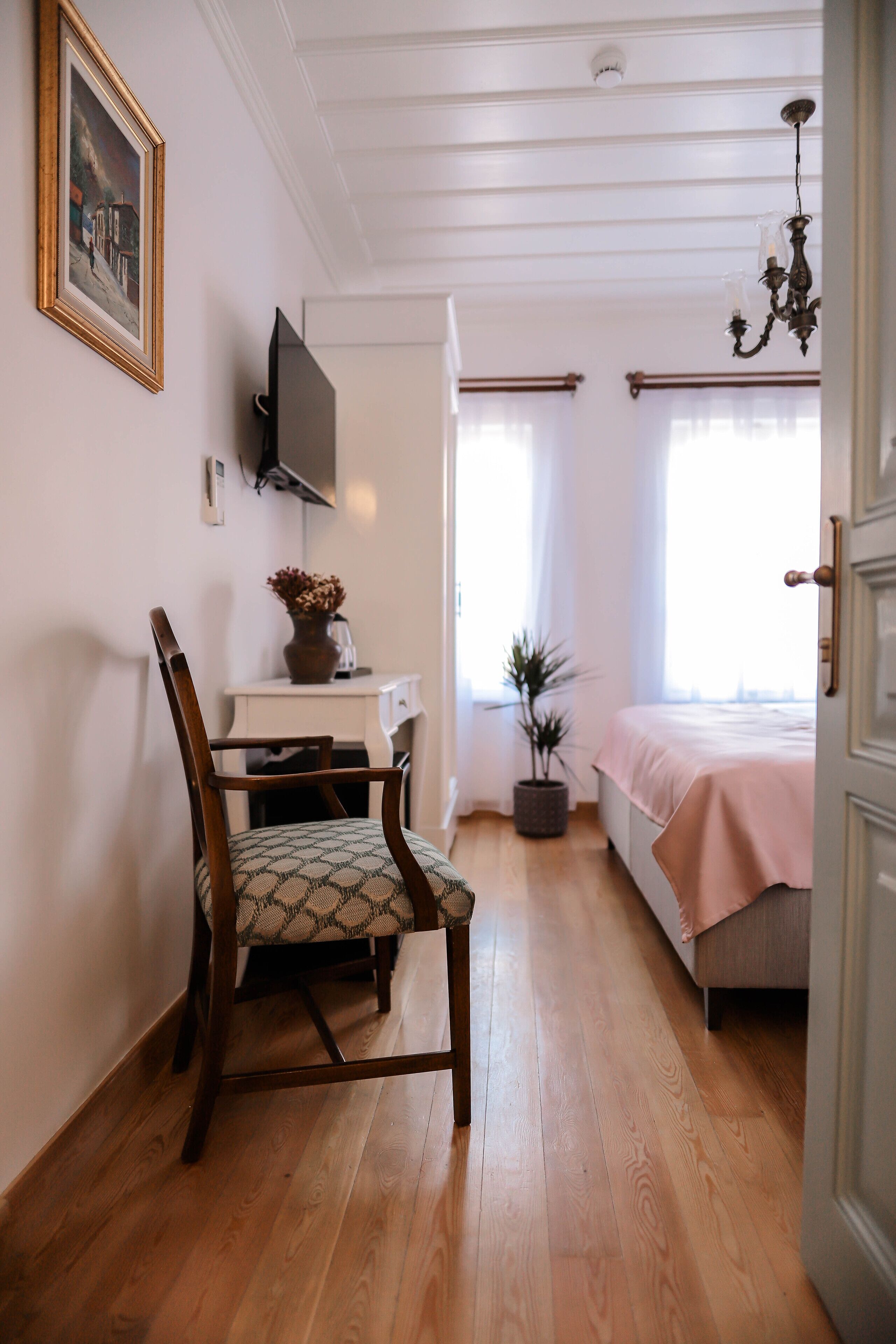 Quarto com piso de madeira, cama com colcha rosa, escrivaninha com computador, cadeira, planta em vaso e lustre.