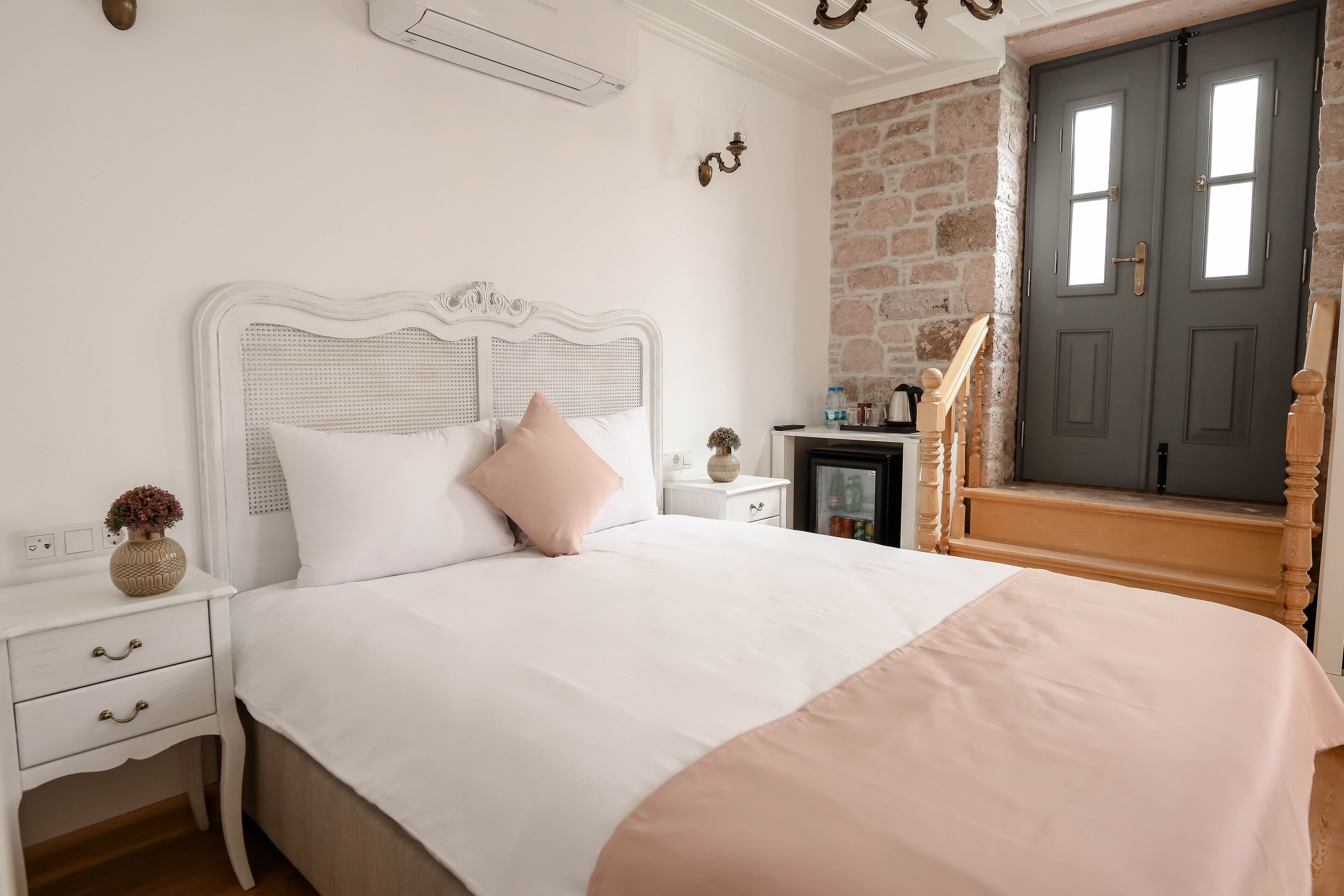 Uma cama bem arrumada com colcha branca e rosa, uma mesa de cabeceira branca com gavetas, um pequeno vaso com flores e uma parede de pedra com escada de madeira.