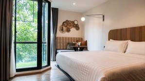 Quarto de hotel moderno com uma cama grande, uma mesa pequena com uma cadeira, um elemento decorativo na parede e uma janela ampla com cortinas.