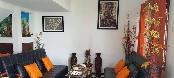 Sala de estar com sofá preto, almofadas laranja e mesa de centro com objetos decorativos. Há quadros nas paredes e um vaso grande com galhos.