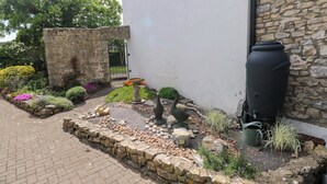 Um jardim com muro de pedra, um bebedouro para pássaros, churrasqueira e diversas plantas.