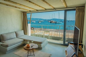 Sala de estar moderna com sofá, uma mesinha e um copo de vinho. Possui uma grande porta de vidro que dá para uma varanda com vista para barcos e o mar.