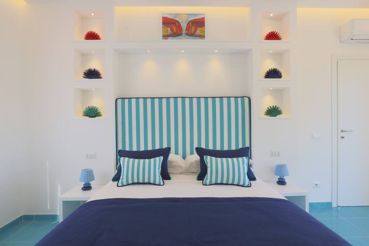 Quarto com cabeceira listrada, duas mesas de cabeceira com luminárias e objetos decorativos nas prateleiras.