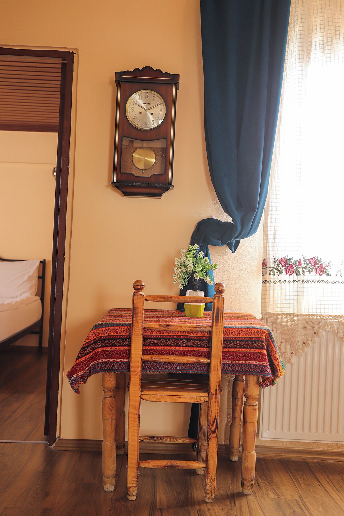 Uma cadeira de madeira com toalha de mesa estampada, um relógio de parede, um vaso com planta e uma janela com cortinas.